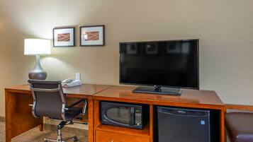Quarto de hotel com secretária de madeira, televisão, micro-ondas, frigorífico, cadeira e candeeiro.