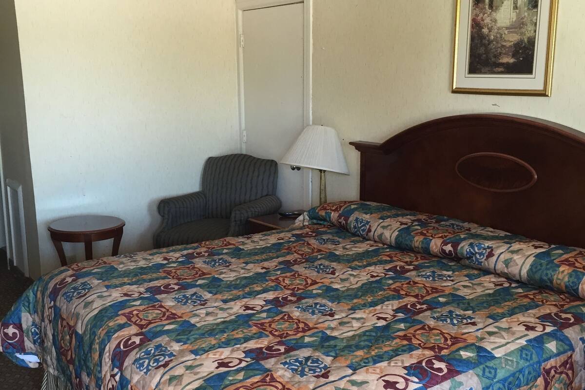 Quarto de hotel com cama grande, uma cadeira, uma mesinha, um abajur e um quadro na parede.