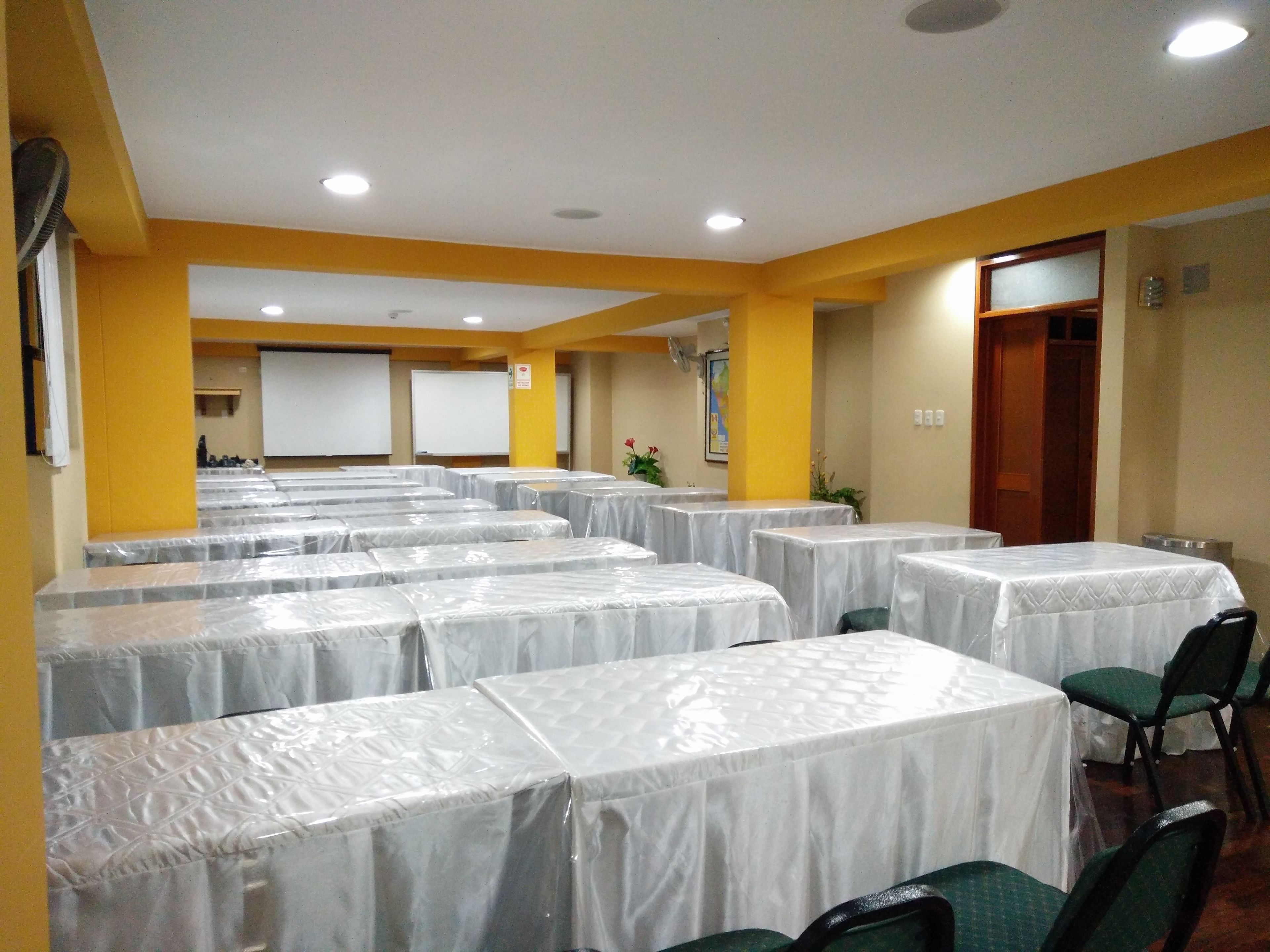 Sala de conferência com mesas cobertas por plástico, equipada com cadeiras, quadro branco e tela de projeção.