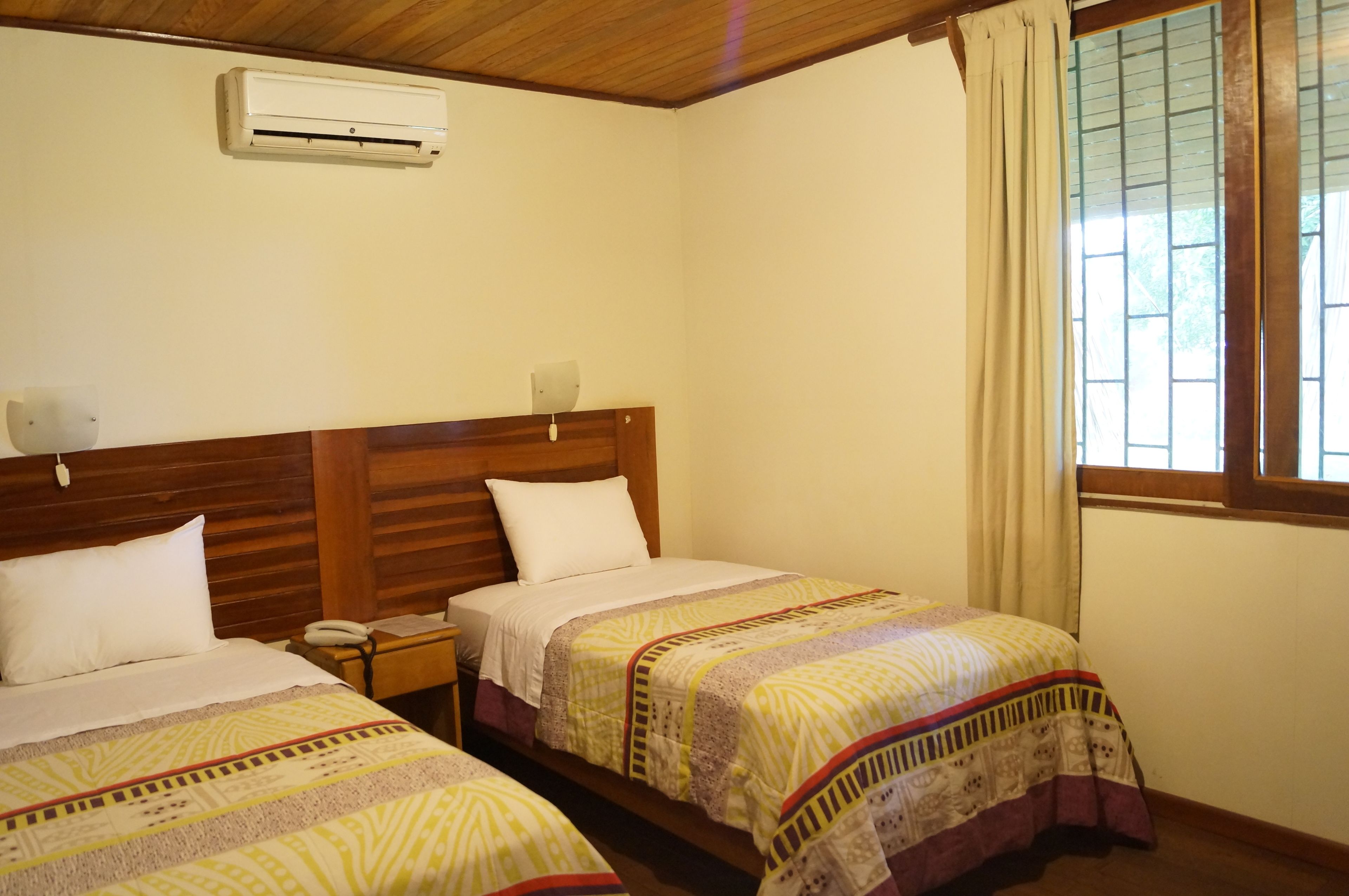 Quarto de hotel com duas camas, cabeceiras de madeira, ar condicionado fixado na parede, mesa de cabeceira com abajur e janela com cortinas.