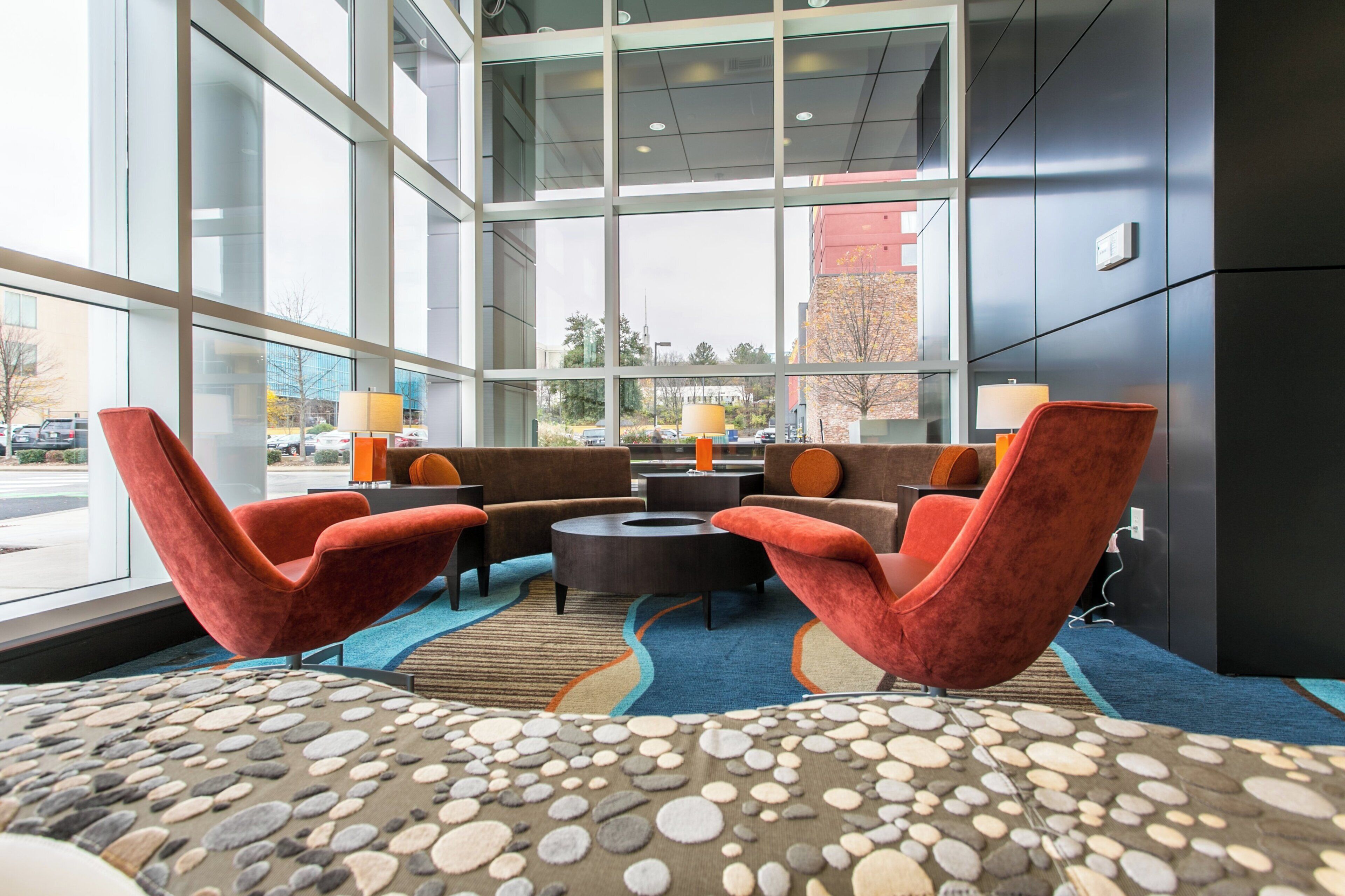 Um lobby moderno com cadeiras vermelhas, um sofá e uma mesa de centro. Grandes janelas proporcionam vista do exterior.
