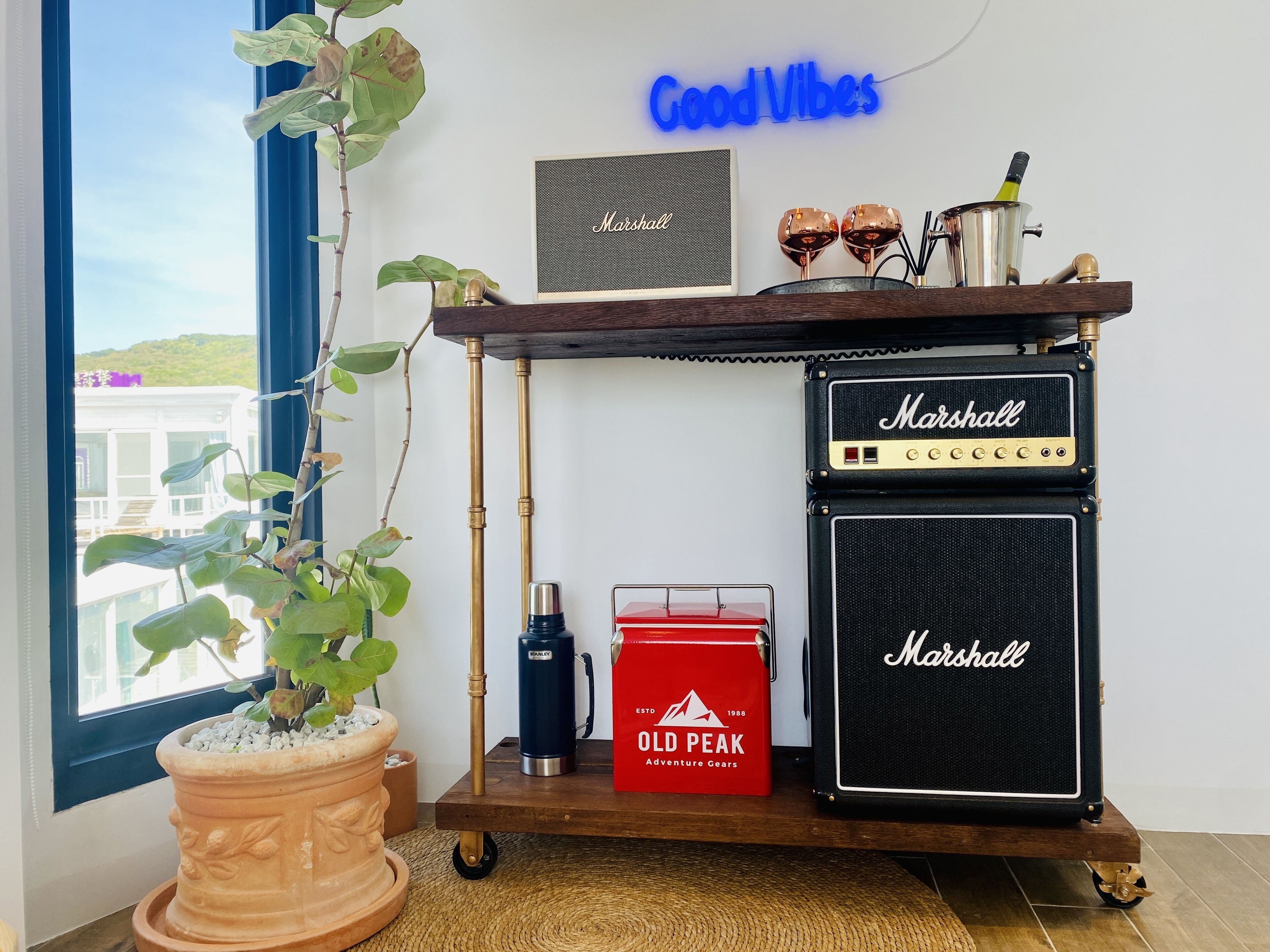 Um quarto com janela, uma planta, um amplificador Marshall, uma caixa vermelha com a inscrição 'Old Peak Adventure Gears' e um letreiro de neon com a mensagem 'Boas Vibrações'.