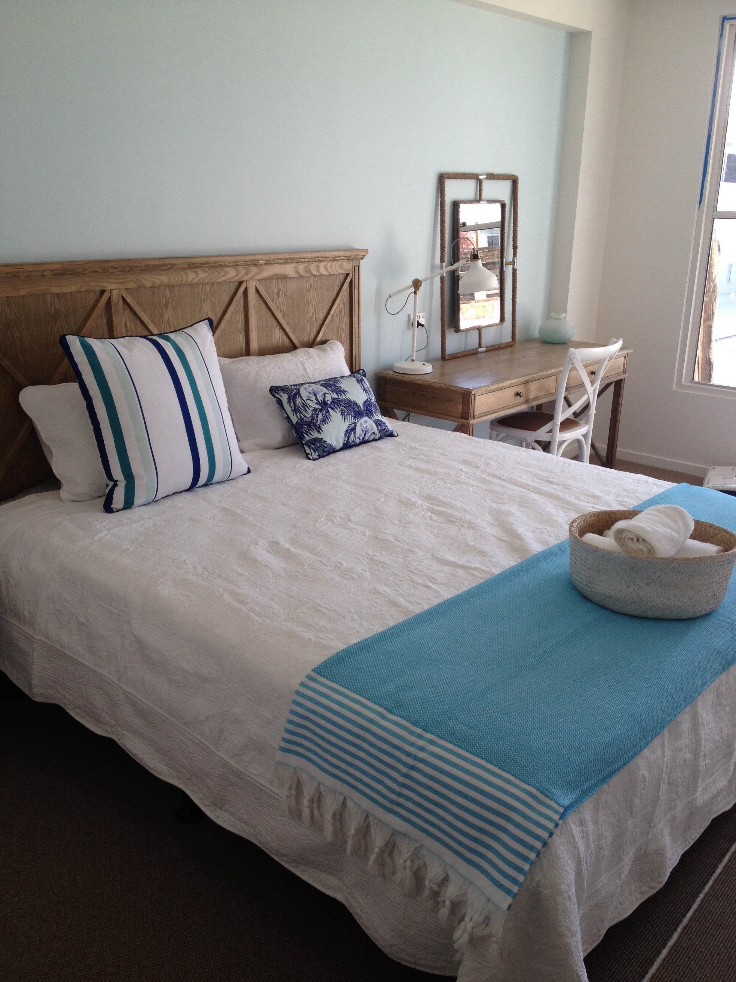 Cama bem arrumada com cabeceira de madeira, dois travesseiros, manta listrada em azul e branco, e uma pequena mesa de cabeceira com abajur e espelho.