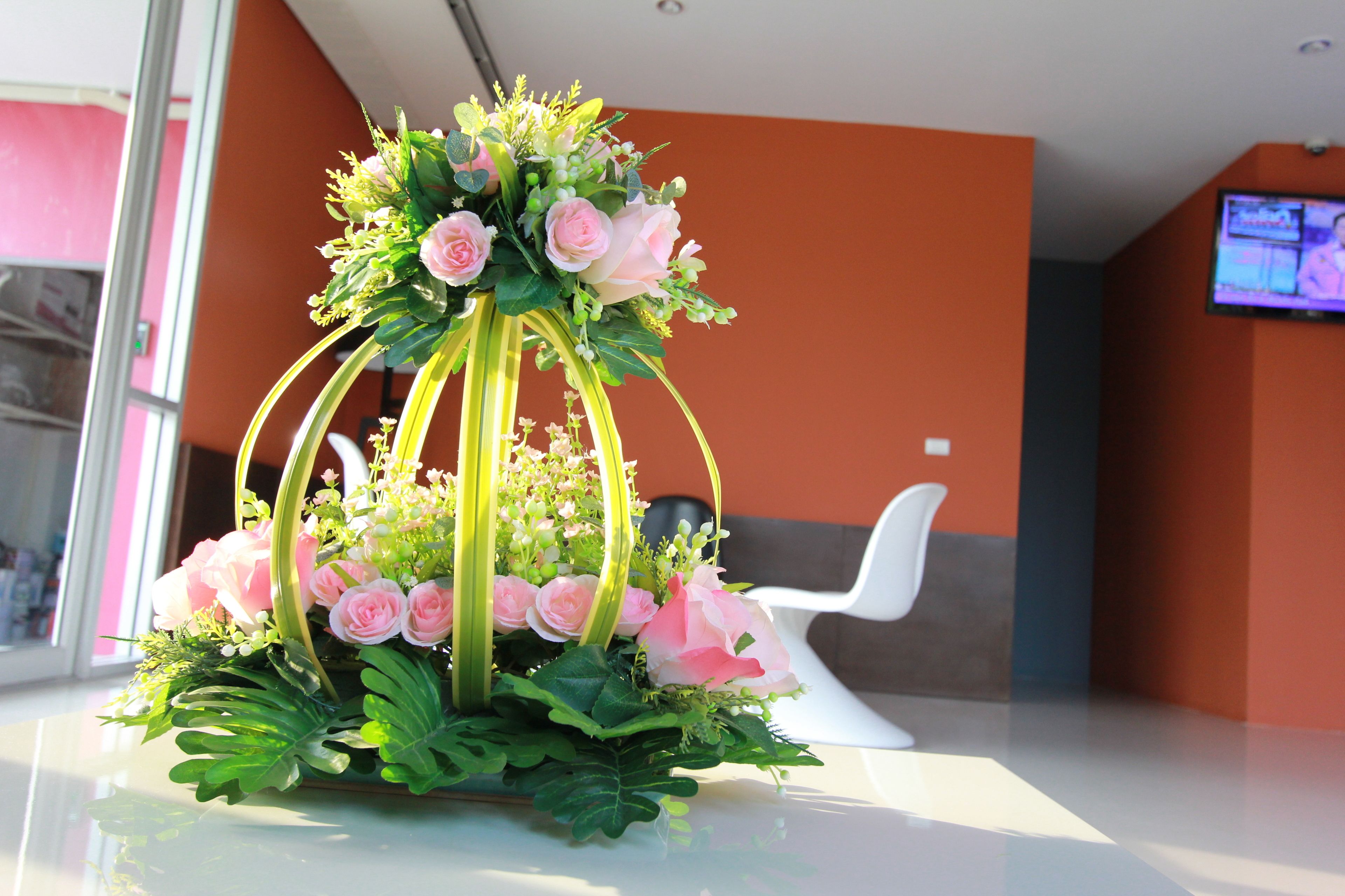 Uma área de recepção com um arranjo floral sobre uma mesa, uma cadeira branca e uma TV fixada na parede.
