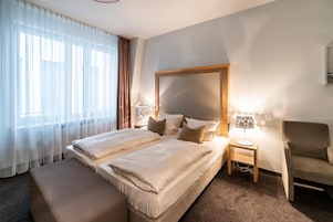 Quarto de hotel com uma cama grande, uma mesa de cabeceira com abajur, uma cadeira e uma janela com cortinas leves.