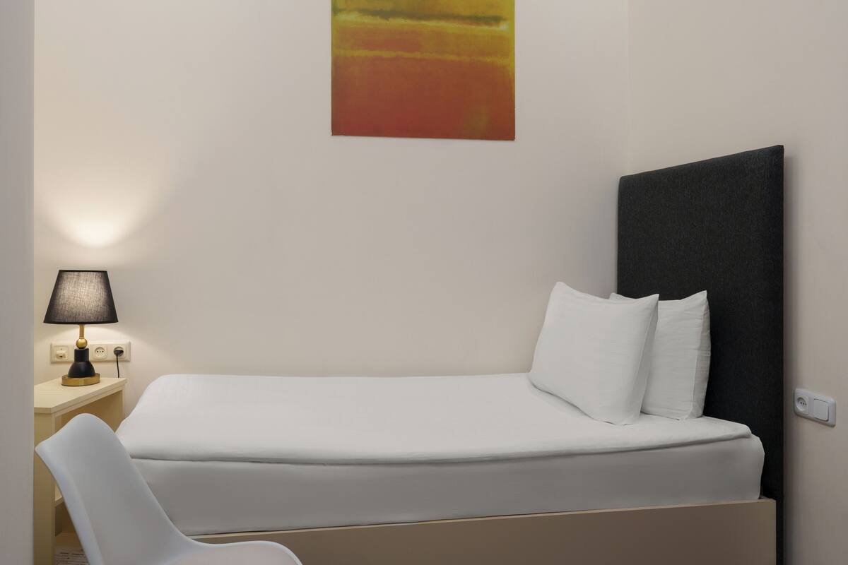 Uma cama de solteiro com roupa de cama branca, uma luminária de cabeceira e um quadro abstrato pendurado na parede.