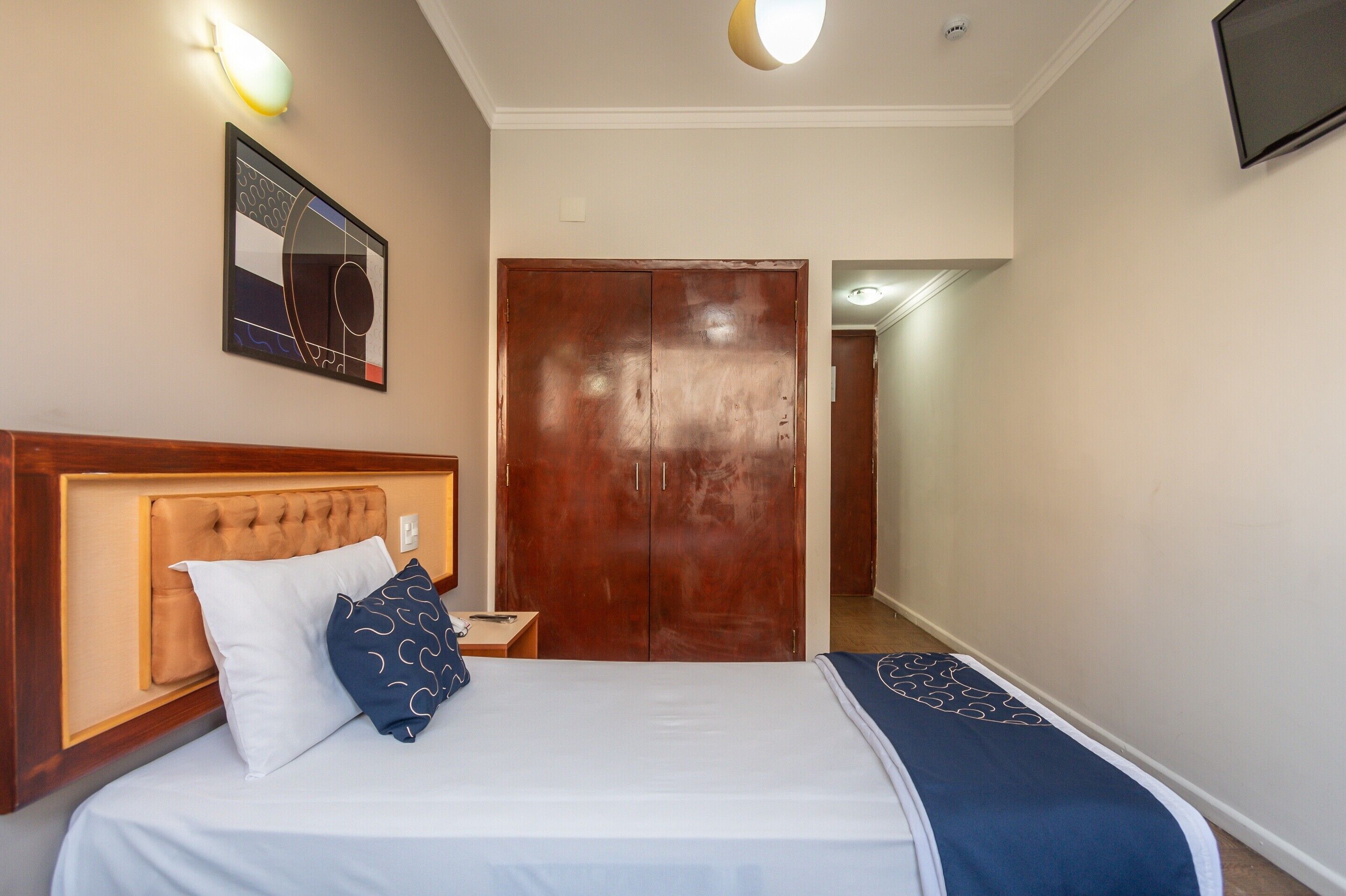 Quarto de hotel com uma cama, um guarda-roupa de madeira, uma mesinha de cabeceira e uma televisão na parede.