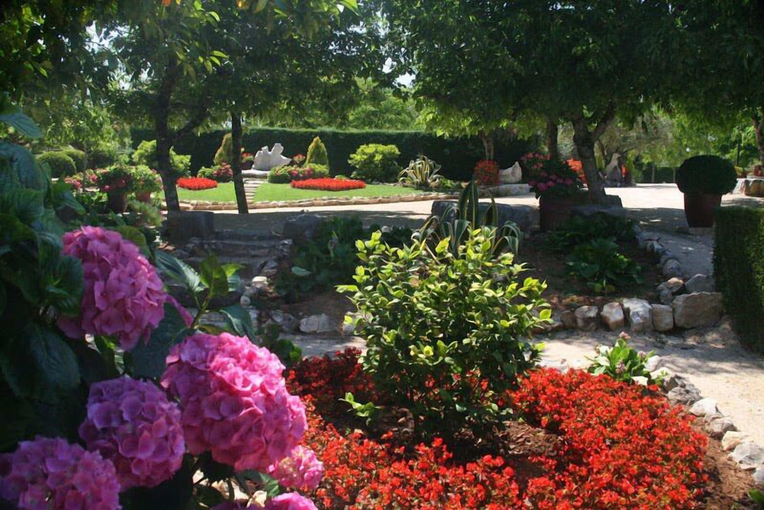 Um jardim bem cuidado, com diversas plantas e flores, um caminho de pedra e sebes bem aparadas.
