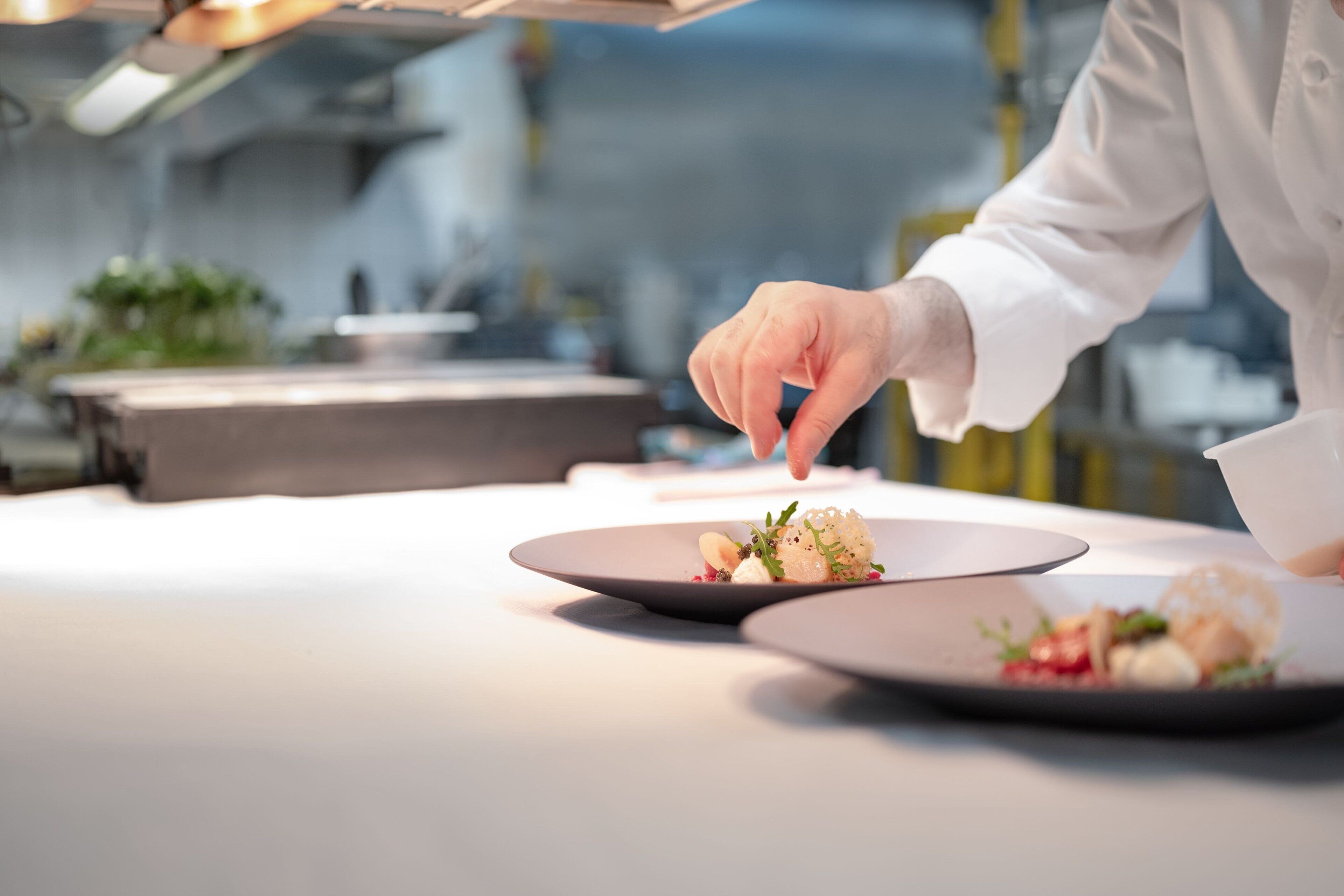Mão de chef segurando uma faca, preparando comida em um prato em uma cozinha.