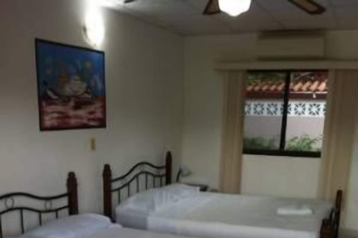 Quarto de hotel com duas camas de solteiro, um ventilador de teto, uma janela com vista e um quadro na parede.