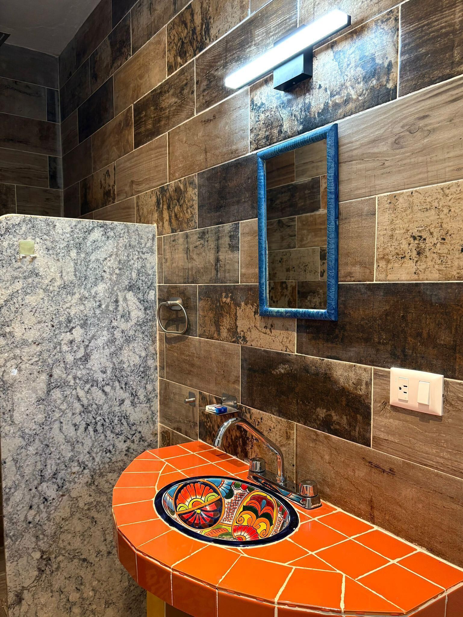 Casa de banho com bancada de granito, lavatório com azulejos coloridos e espelho numa parede de madeira.