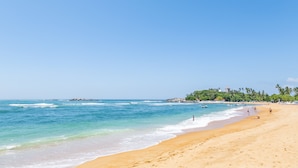 Uma praia com águas azul-claras, areal arenoso e algumas pessoas caminhando.