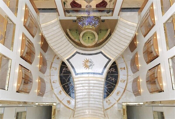 Um amplo átrio com escadaria central, lustre e teto decorado com signos do zodíaco.