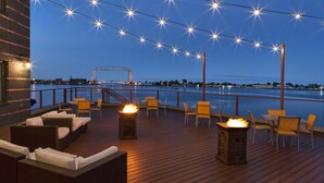 Um terraço com área de estar ao ar livre, lareira e luzes de corda, com vista para um cais e uma ponte.