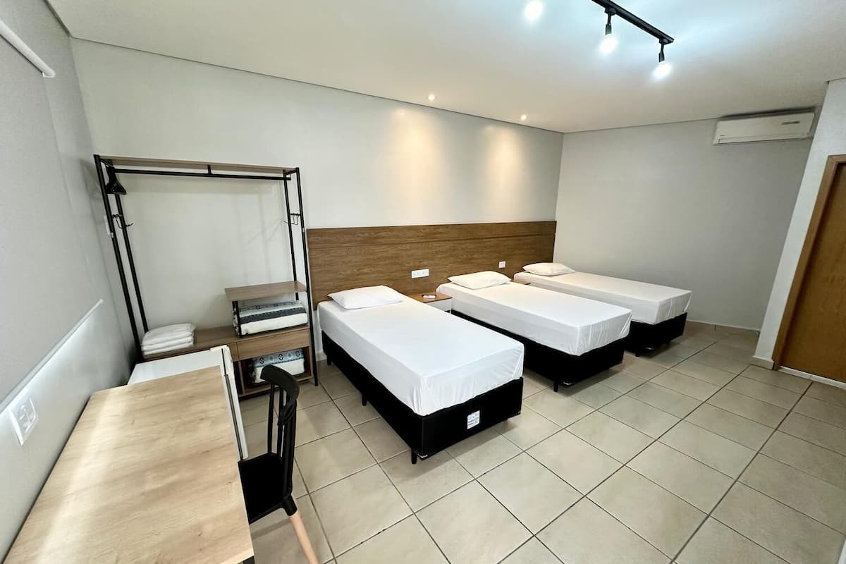 Quarto de hotel com três camas de solteiro, uma escrivaninha, uma cadeira, um guarda-roupa e um espelho.