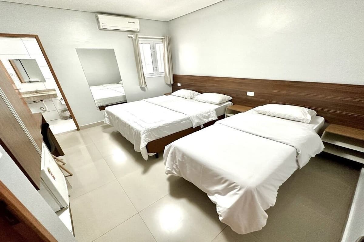 Quarto de hotel com duas camas de solteiro, cabeceira de madeira, mesinha de cabeceira e espelho.