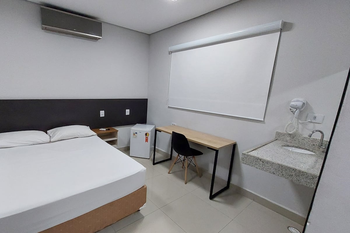 Quarto de hotel com cama, escrivaninha, pequeno frigorífico, secador de cabelo e lavatório.