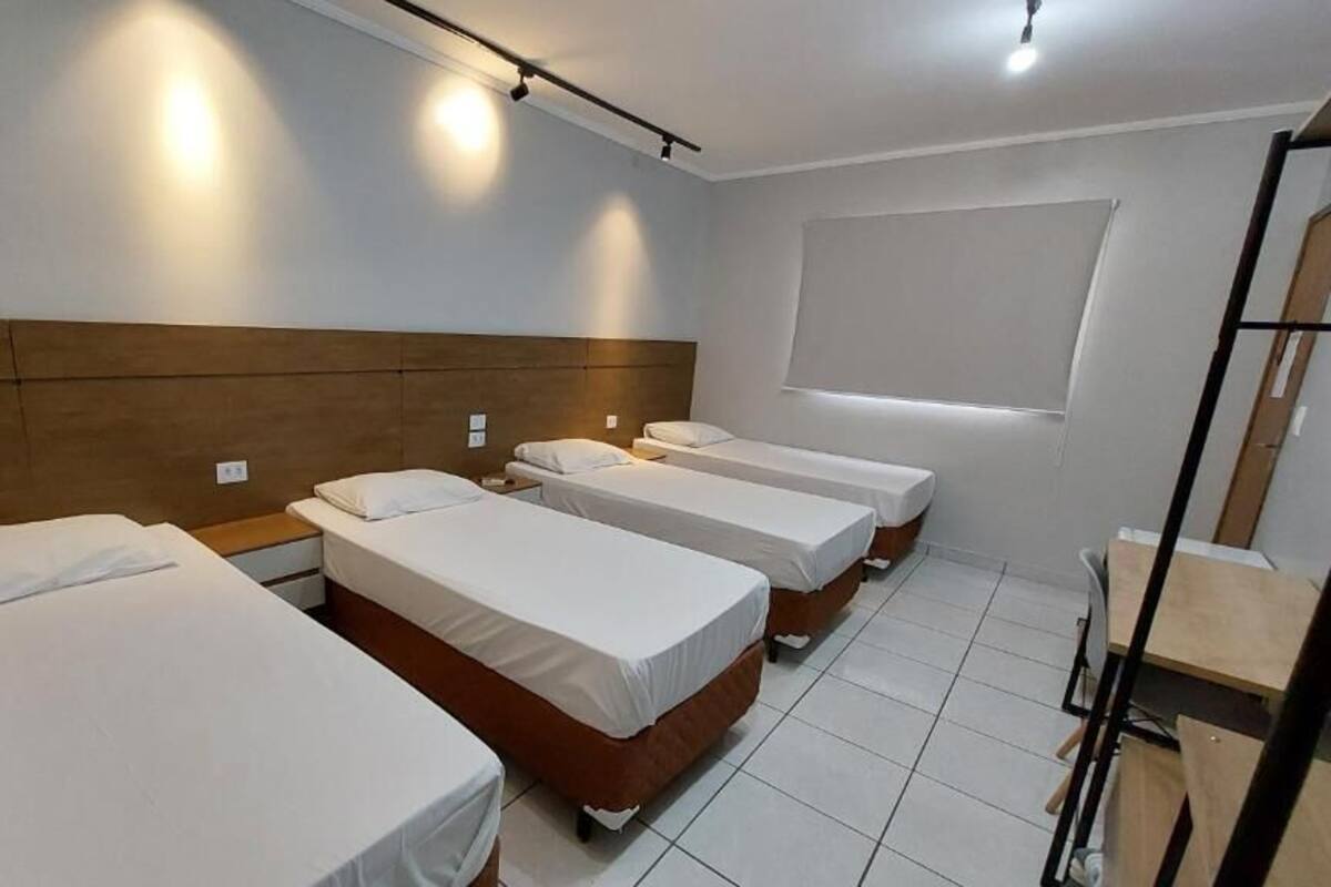 Quarto de hotel com quatro camas de solteiro, cabeceira de madeira, uma janela grande e uma escrivaninha com cadeira.