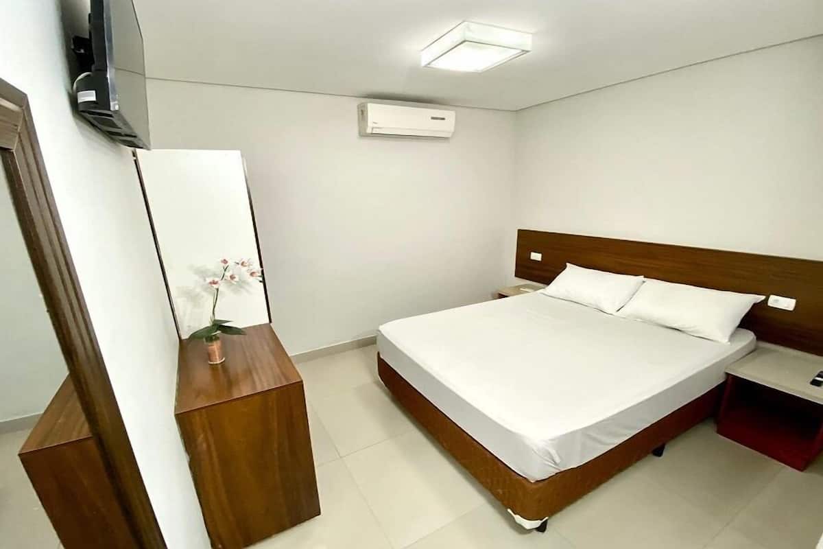 Quarto de hotel com uma cama, uma mesa de cabeceira com um vaso de flores, um espelho, uma TV fixada na parede e um aparelho de ar condicionado.