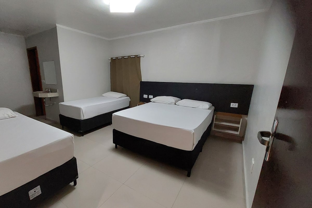Quarto de hotel com duas camas de solteiro, uma pequena mesa de cabeceira, um abanador, um espelho e uma janela com cortinas.