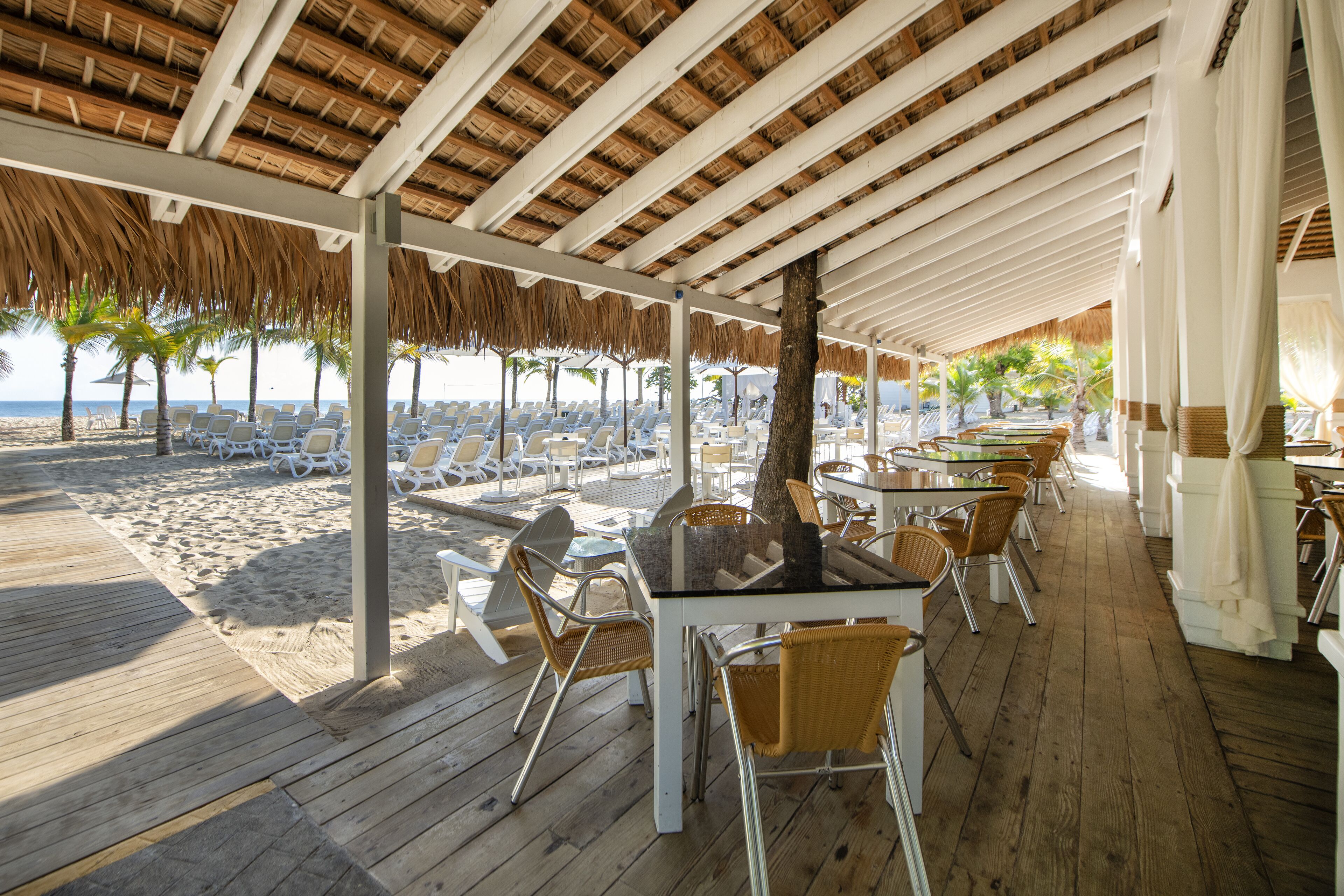 Um restaurante à beira-mar com mesas ao ar livre, um deque de madeira e um telhado de palha.