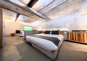 Quarto moderno com uma cama grande, vigas de madeira no teto, uma televisão e uma pequena escrivaninha.