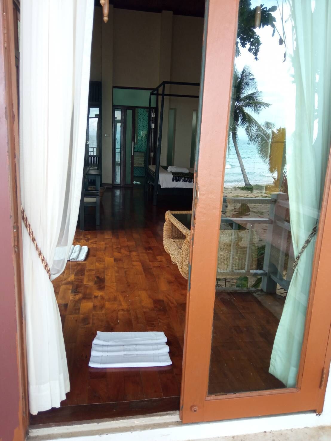 Quarto com piso de madeira, cama com colcha branca, cadeira de vime e vista da praia através de uma porta de vidro.
