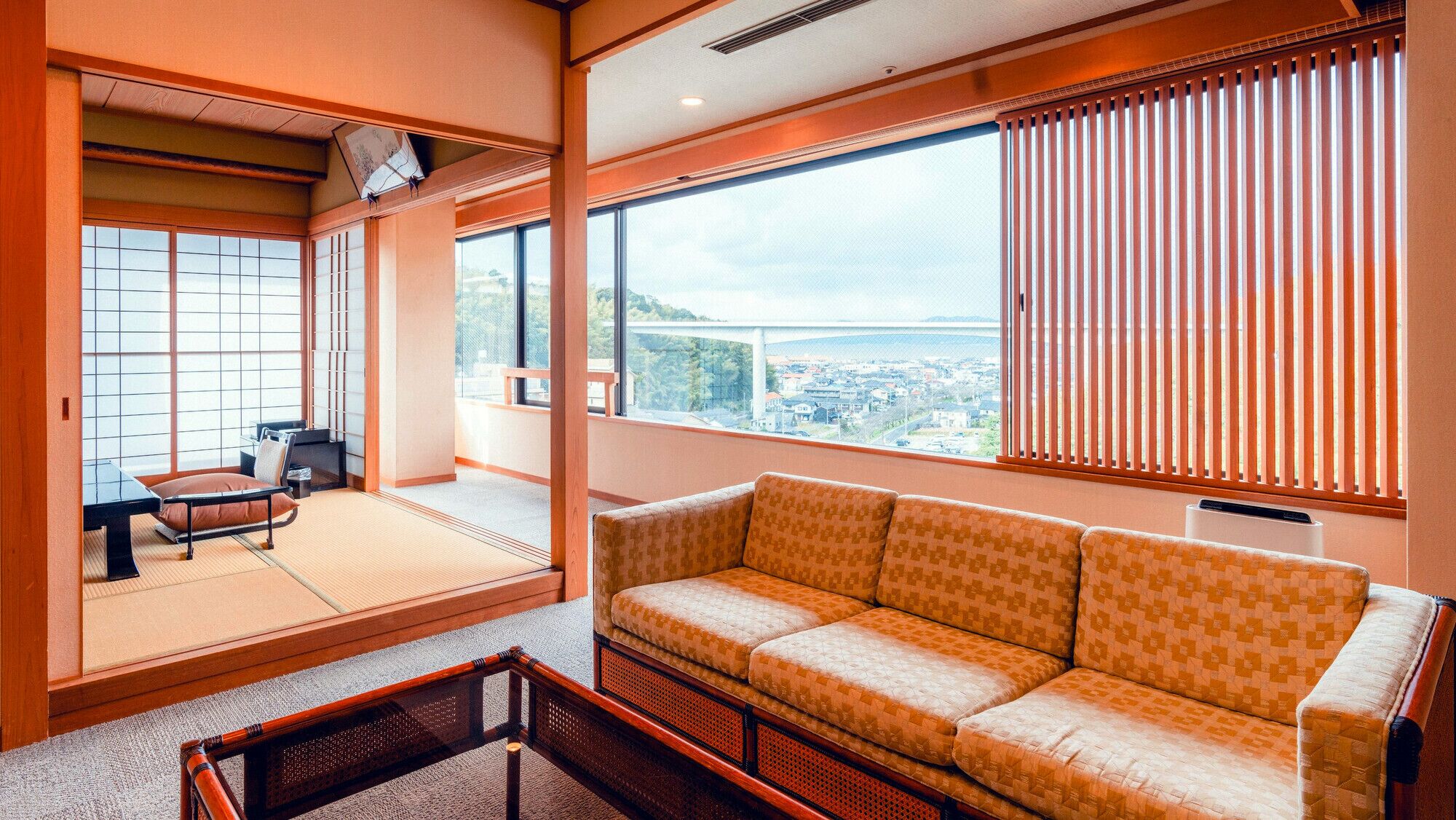 Uma sala de estar moderna com piso de tatame, um sofá, uma mesinha de centro e vista para um horizonte urbano através de grandes janelas.
