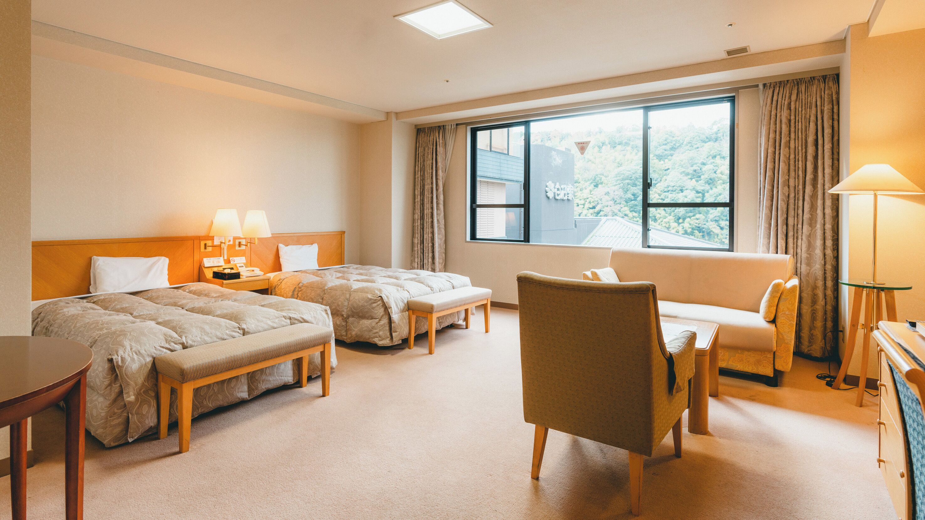 Quarto de hotel com duas camas, uma escrivaninha, uma cadeira, um sofá, um abajur e uma janela com vista.