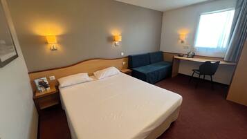 Quarto de hotel com cama, escrivaninha, cadeira e duas luminárias fixadas na parede.