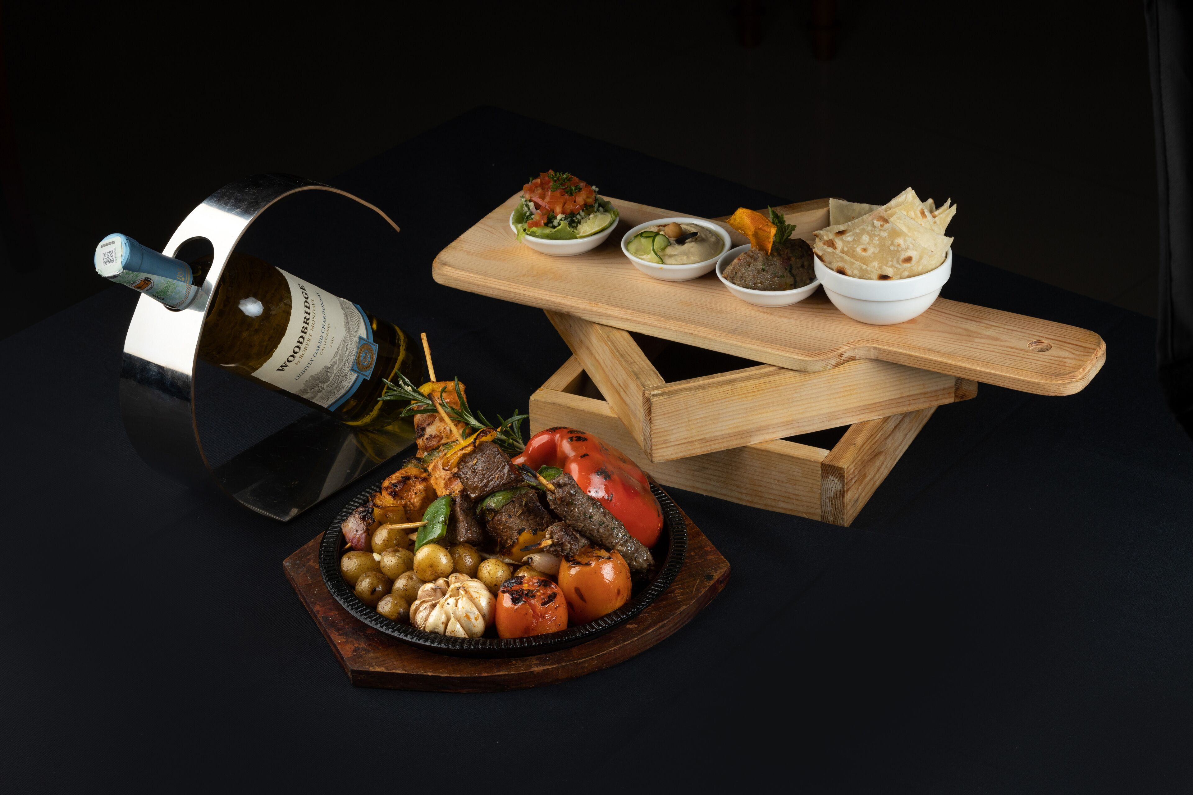 Uma mesa de madeira com uma garrafa de vinho, uma saladeira e um prato de legumes grelhados.