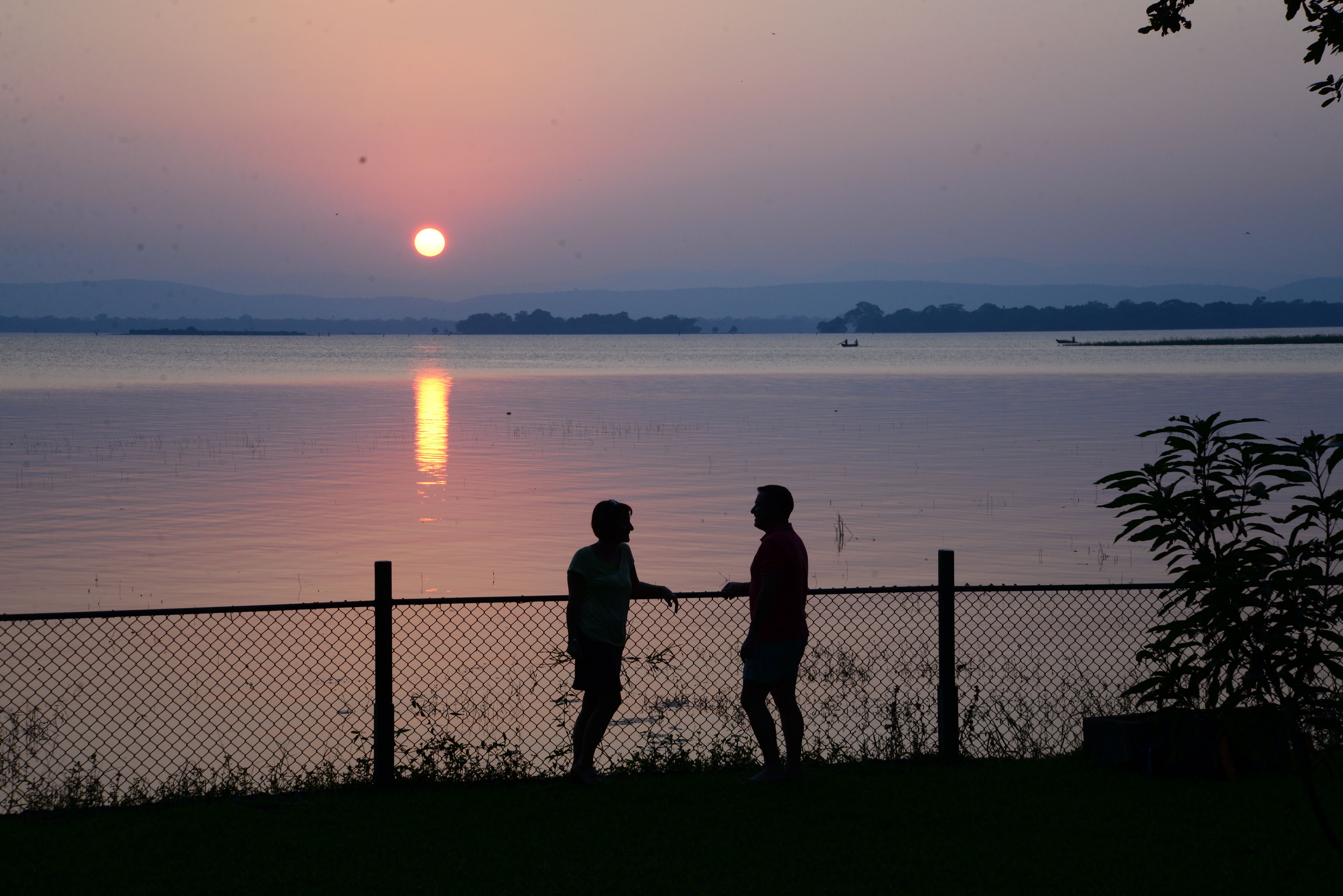 Duas pessoas perto de uma cerca, observando um corpo d'água calmo ao pôr do sol.