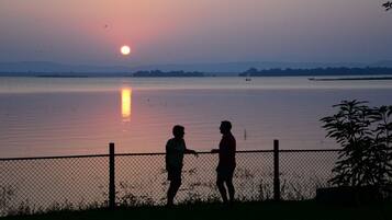 Duas pessoas perto de uma cerca, observando um corpo d'água calmo ao pôr do sol.