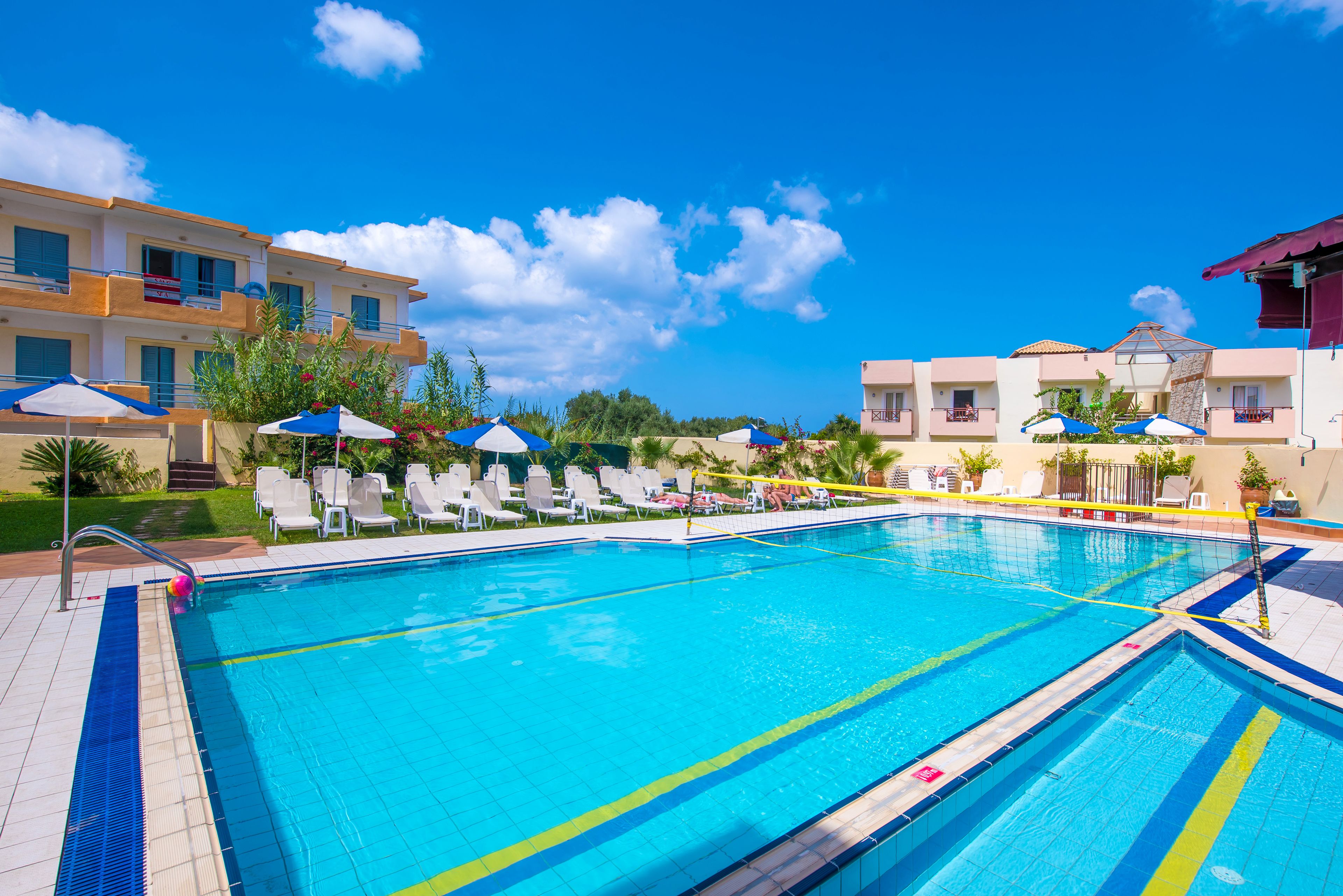 Piscina com espreguiçadeiras e guarda-sóis, cercada por edifícios com varandas e um céu azul límpido.