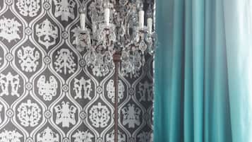 Uma cadeira estofada preta com encosto curvo e pés de madeira, posicionada diante de um papel de parede estampado e um lustre de cristal alto e ornamentado.