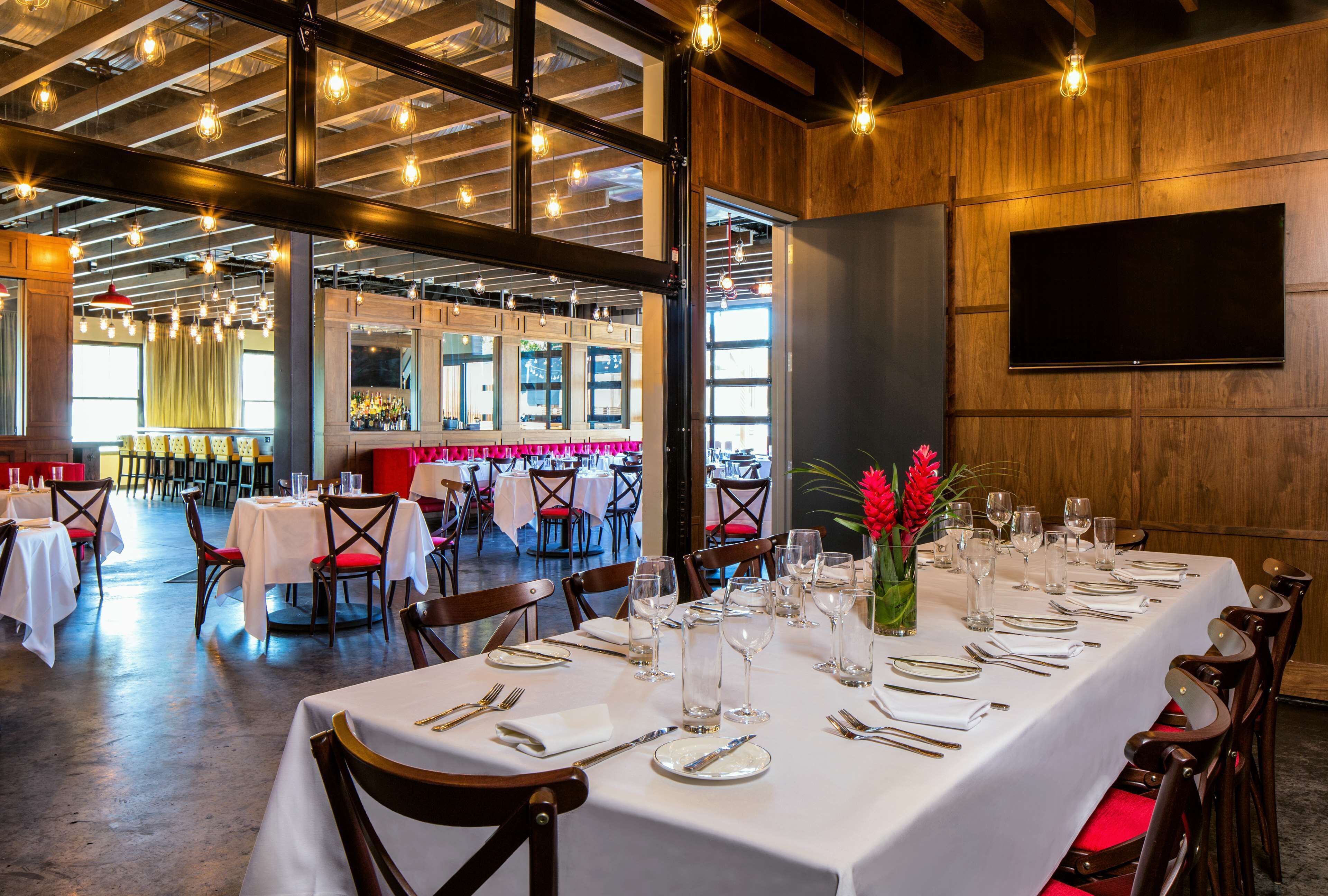Uma sala de jantar com uma mesa comprida posta para refeição, vigas de madeira no teto e uma televisão grande fixada na parede.