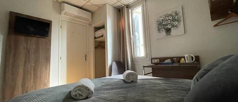 Quarto de hotel com uma cama, escrivaninha, cadeira, televisão e janela com cortinas.
