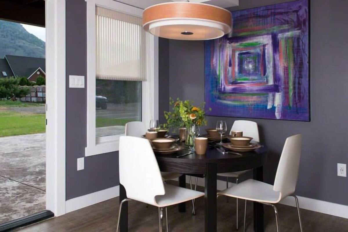 Uma sala de jantar com mesa redonda posta para quatro lugares, cadeiras brancas, um vaso com flores e uma pintura abstrata colorida na parede.