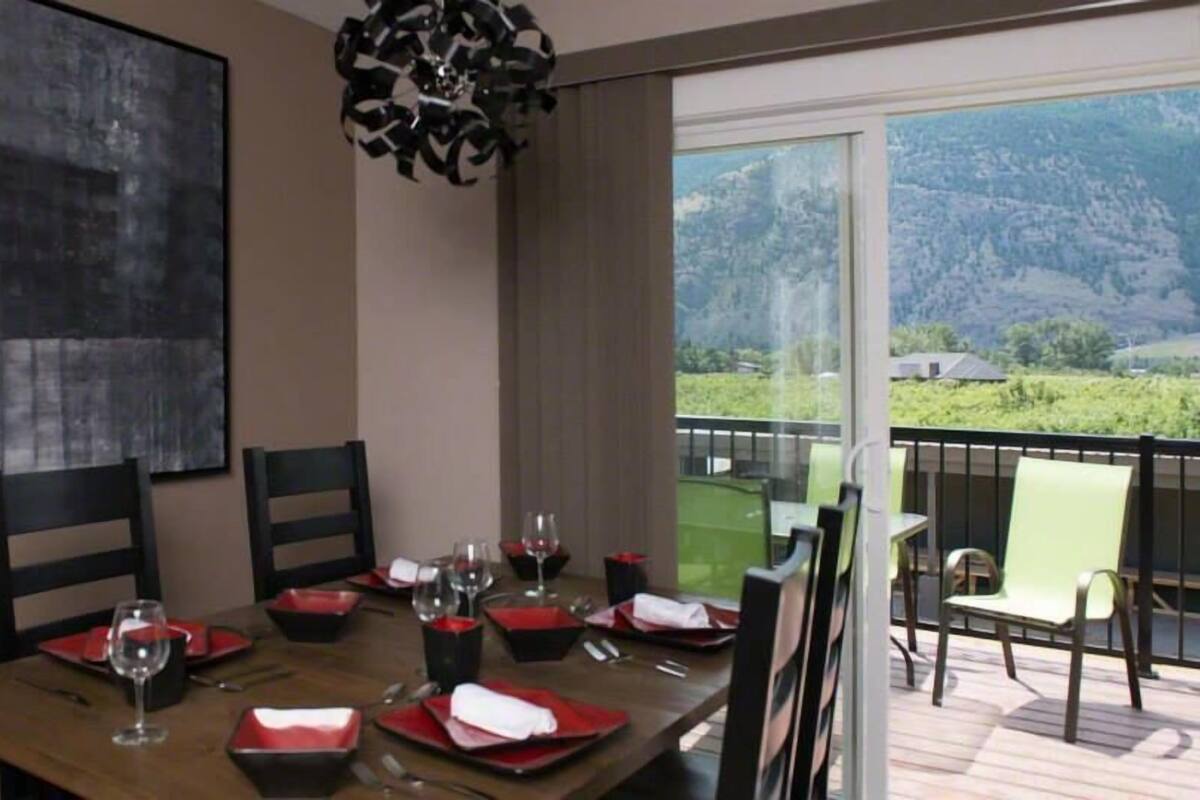 Uma área de refeições com uma mesa posta para dois, com louças vermelhas e pretas, e vista para uma paisagem montanhosa através de uma grande janela.