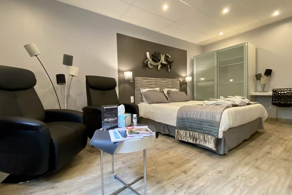 Quarto de hotel moderno com uma cama, duas poltronas, uma mesinha de centro com uma revista, uma TV na parede e um espelho grande.
