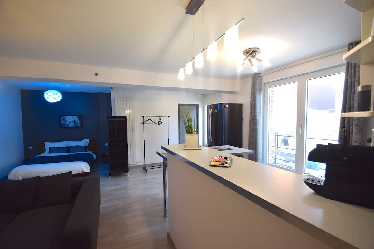 Quarto de hotel moderno com cama, sofá, cozinha compacta e varanda com vista.