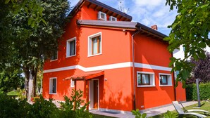 Uma casa vermelha de dois andares com detalhes brancos, uma antena parabólica visível e um jardim bem cuidado com arbustos verdes e gramado.