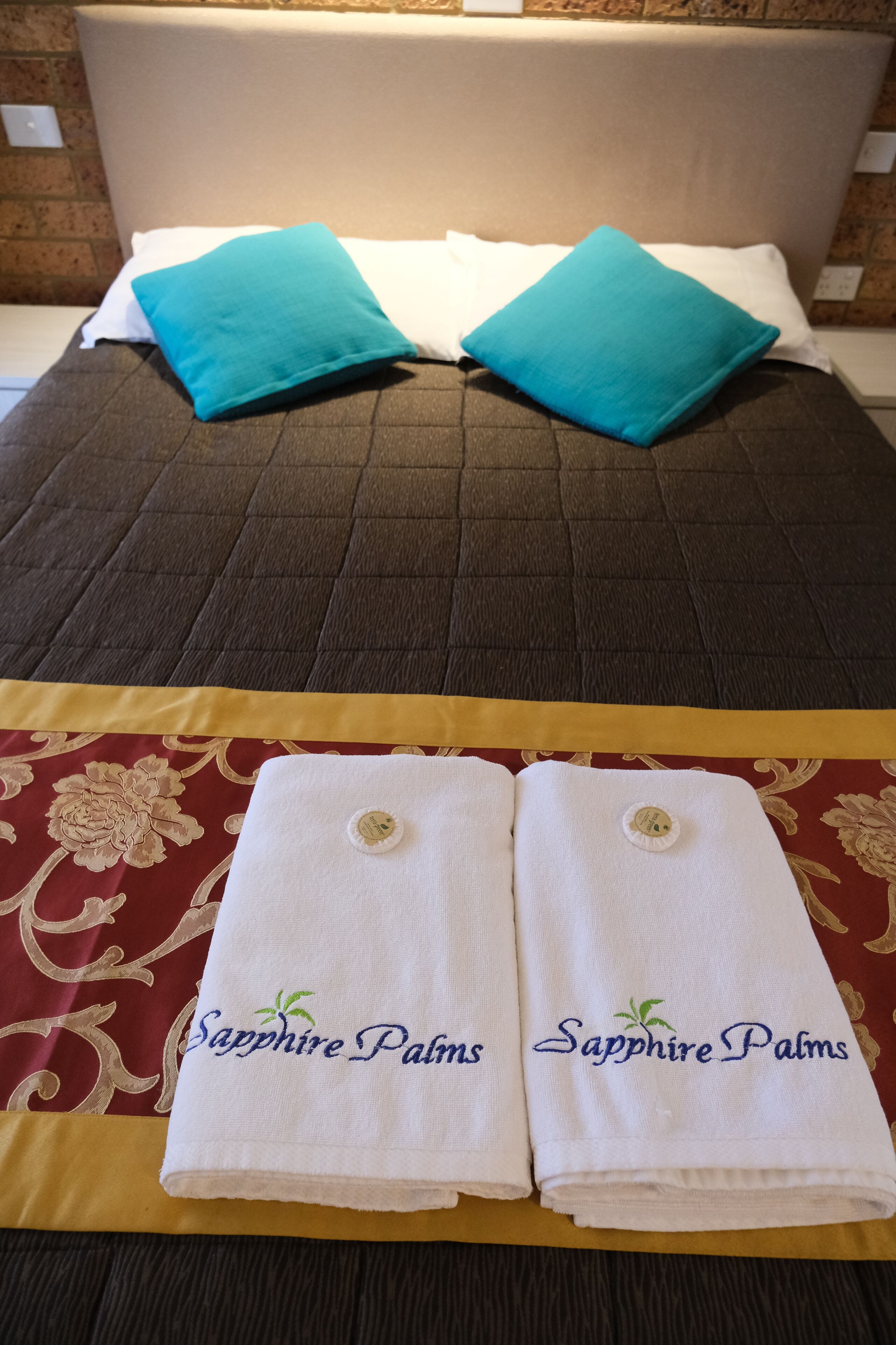 Cama bem arrumada com duas almofadas azul-turquesa e duas toalhas dobradas com o bordado 'Sapphire Palms'.