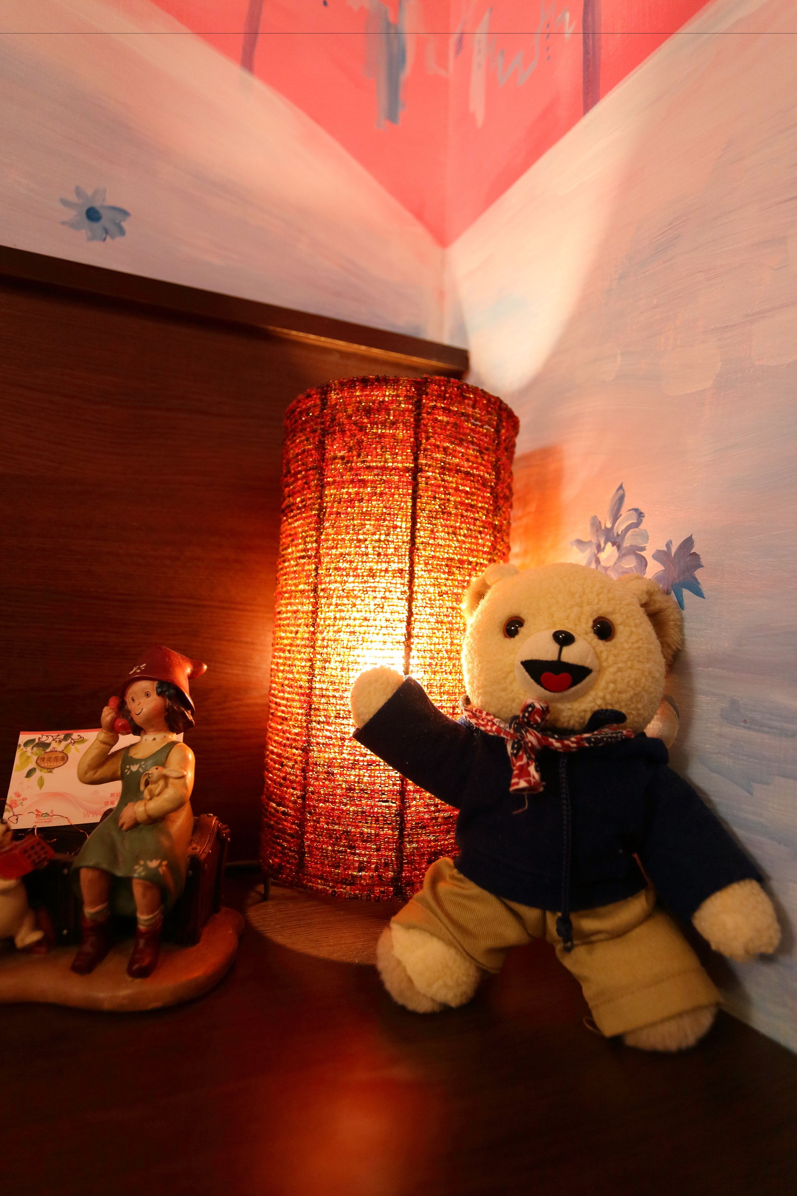 Um urso de pelúcia e uma figurina sobre uma superfície de madeira, com uma luminária vermelha texturizada ao fundo.