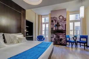 Quarto de hotel com uma cama, uma cadeira azul, uma mesinha com um vaso e uma janela grande com cortinas.