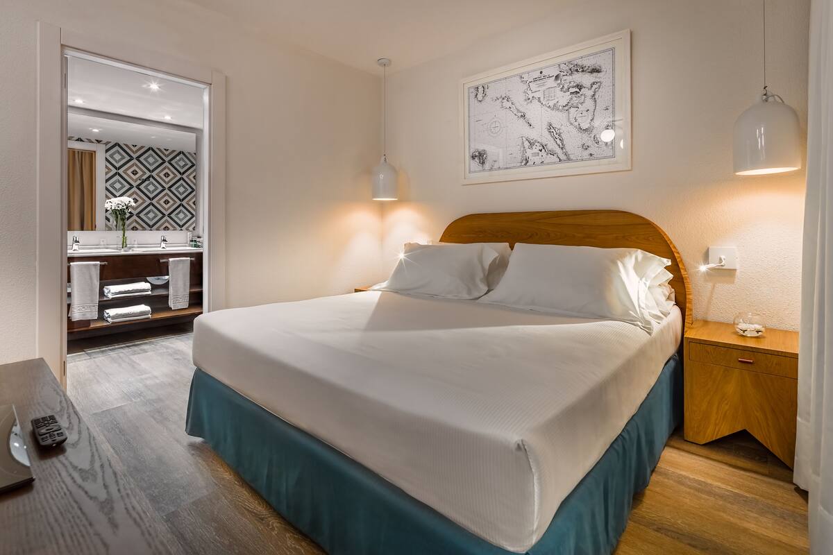 Quarto de hotel bem arrumado com uma cama grande, mesa de cabeceira de madeira, um aparador, um espelho e banheiro com pia e porta-toalhas.