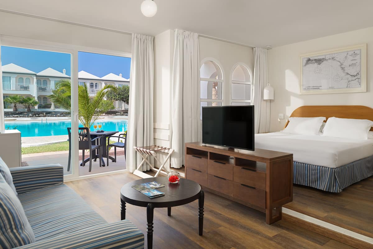 Quarto de hotel com cama, televisão, mesa pequena, sofá e vista para piscina e edifícios.