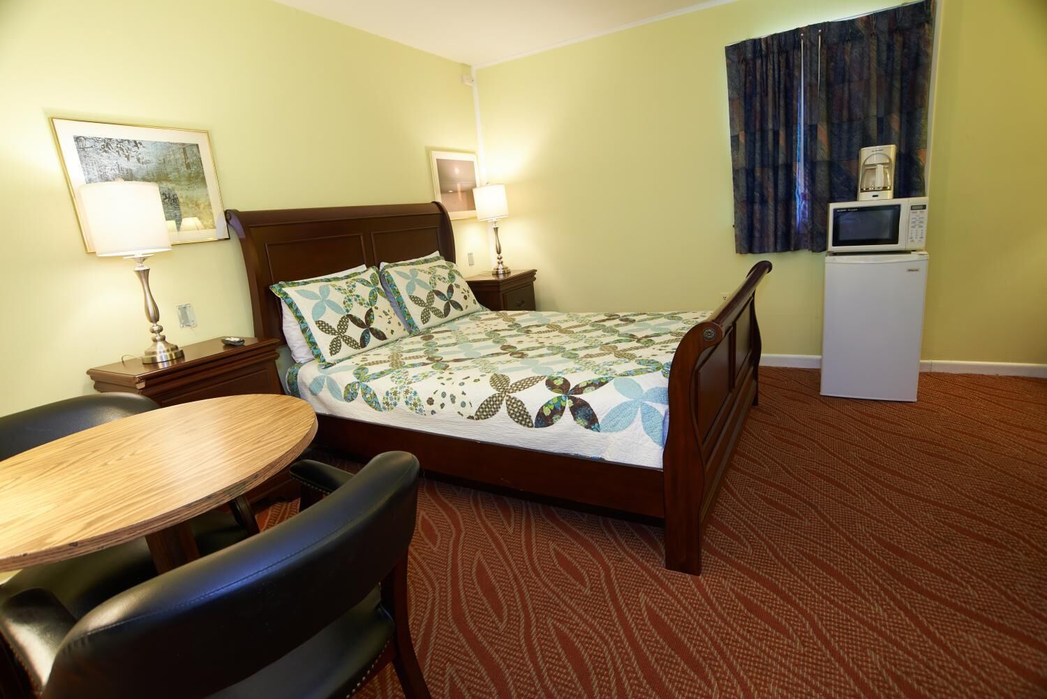 Quarto de hotel com cama, escrivaninha, cadeira, mesa, luminária, micro-ondas e cortina.