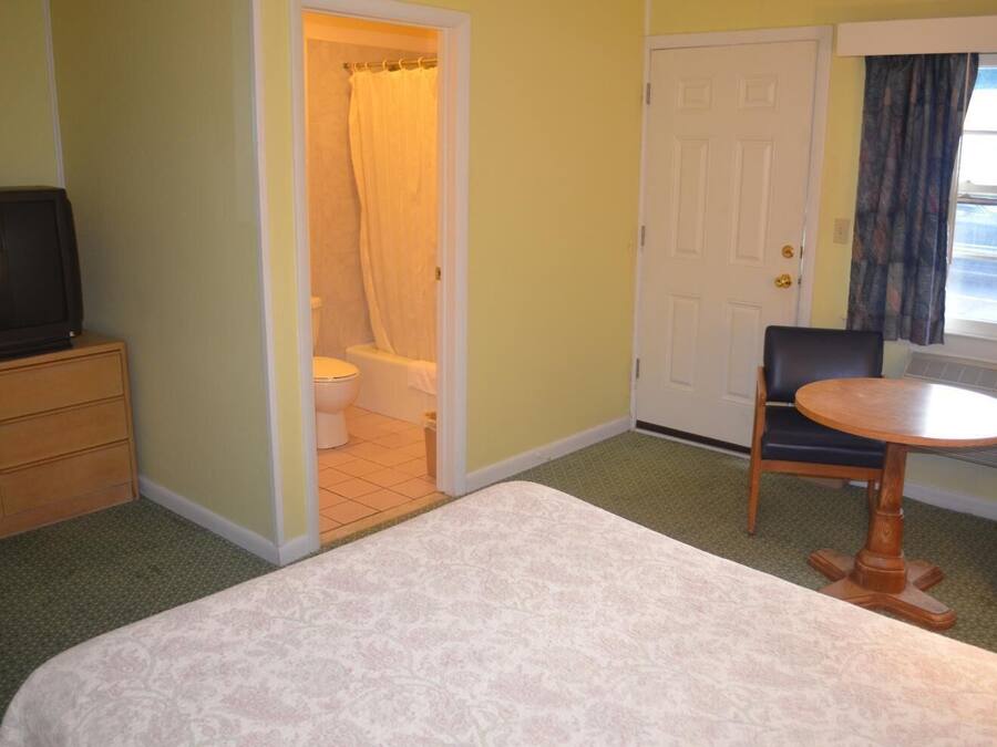 Quarto de hotel com cama, uma pequena mesa, uma cadeira, televisão e banheiro com cortina de chuveiro.