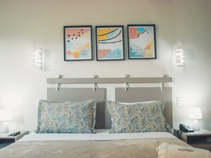 Quarto de hotel com uma cama, duas almofadas, mesa de cabeceira com abajur, telefone e três quadros abstratos na parede.